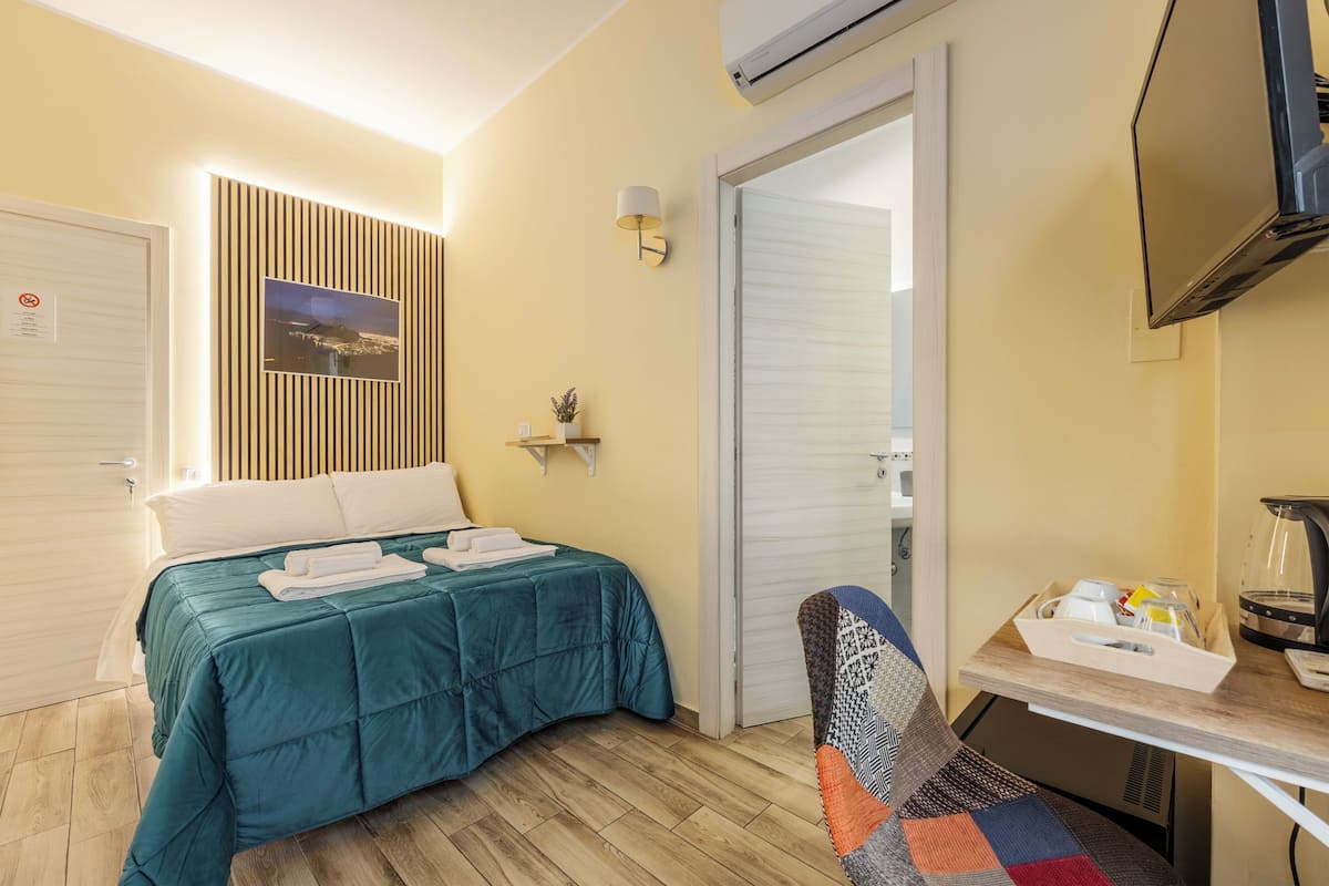 Quarto de hotel com uma cama, uma escrivaninha com cafeteira, uma televisão e uma porta que dá para outro quarto.