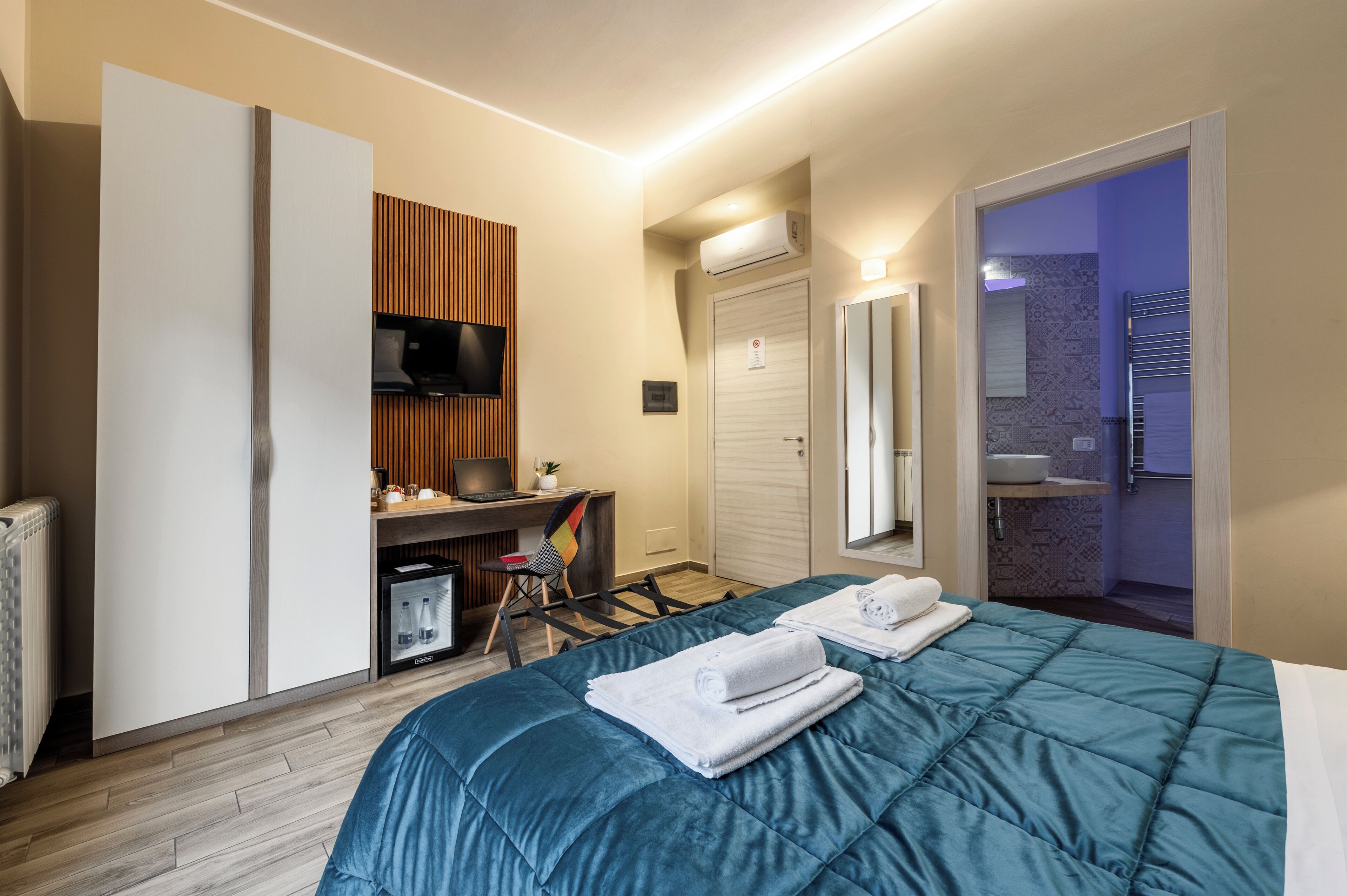 Quarto de hotel moderno com cama, escrivaninha, cadeira, televisão e banheiro visível por uma porta aberta.