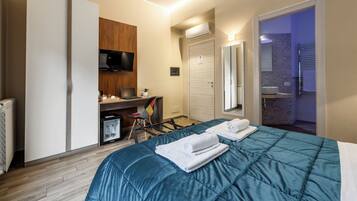 Quarto de hotel moderno com cama, escrivaninha, cadeira, televisão e banheiro visível por uma porta aberta.