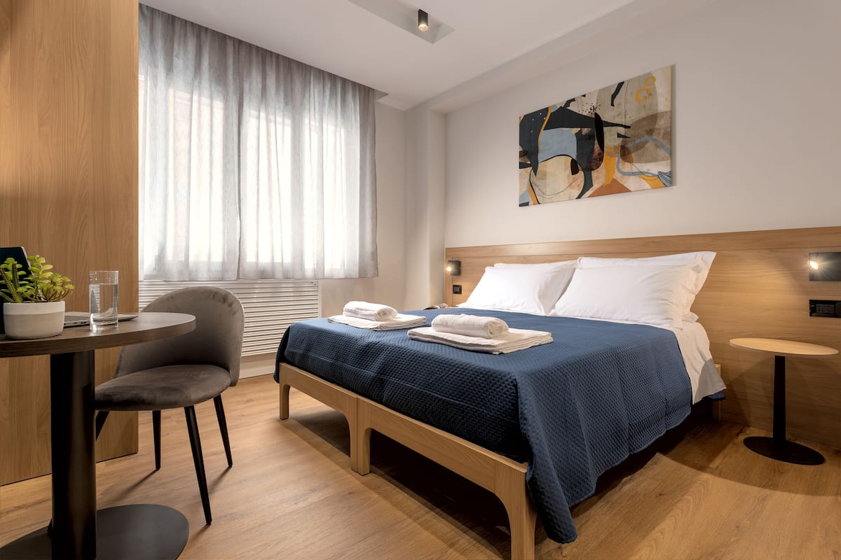 Quarto de hotel com cama, escrivaninha com cadeira, planta em vaso e janela com cortinas.