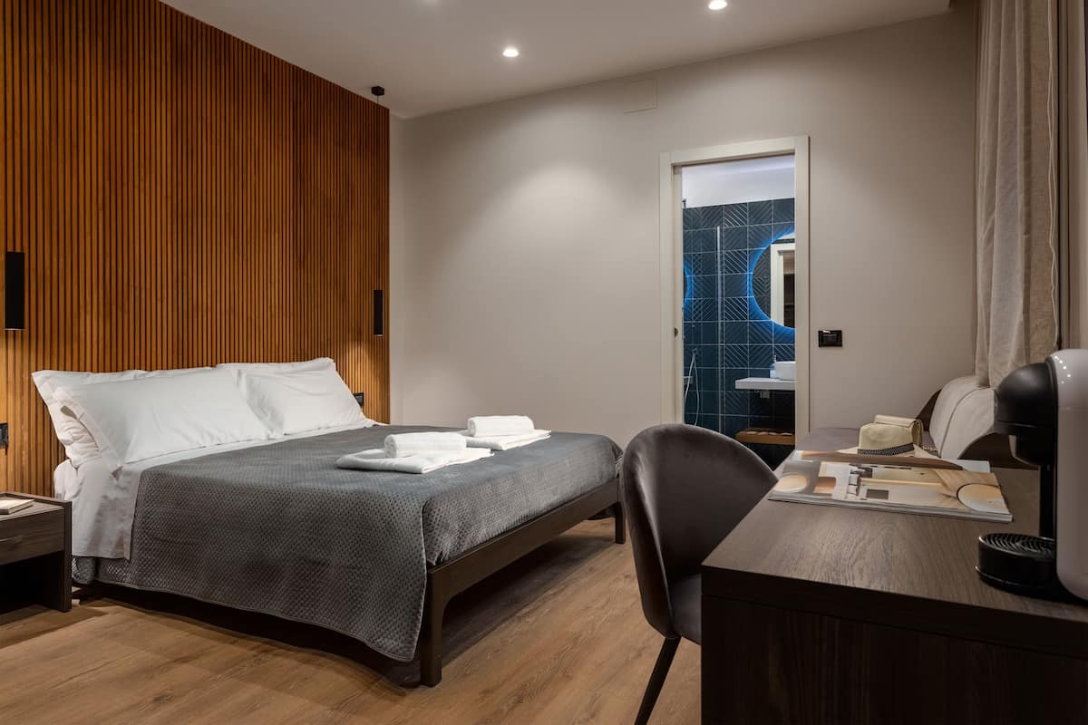 Quarto de hotel moderno com parede de painéis de madeira, cama com roupa de cama branca, escrivaninha com cadeira e banheiro visível por uma porta de vidro.