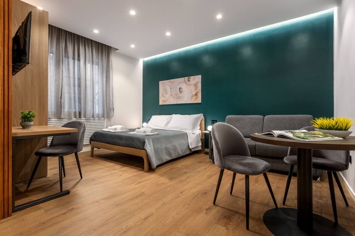 Quarto de hotel moderno com cama, escrivaninha com cadeira, sofá e mesa de jantar.