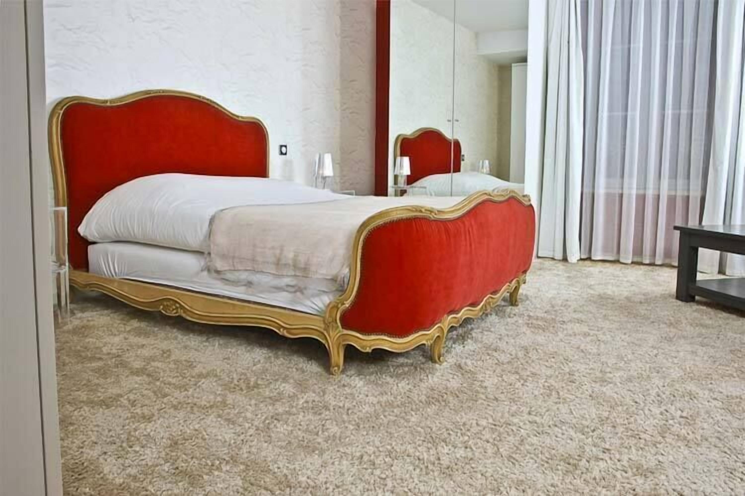 Uma cama com cabeceira vermelha e detalhes dourados, travesseiro branco e manta bege.