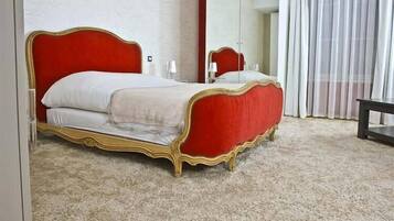 Uma cama com cabeceira vermelha e detalhes dourados, travesseiro branco e manta bege.