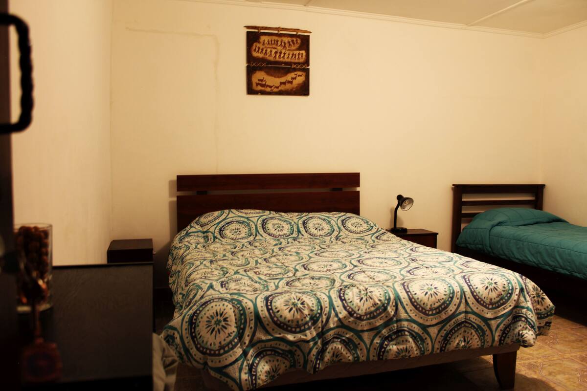 Quarto com cama, mesa de cabeceira, abajur e placa na parede.