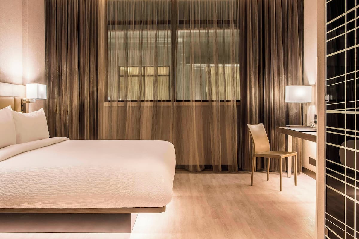 Quarto de hotel com uma cama grande, uma escrivaninha com cadeira e uma janela com cortinas.