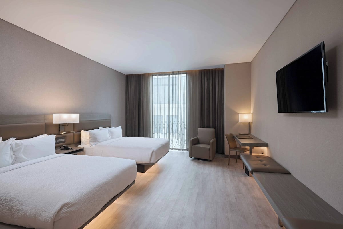 Quarto de hotel com duas camas, uma escrivaninha, uma cadeira, uma televisão e uma janela grande com cortinas.