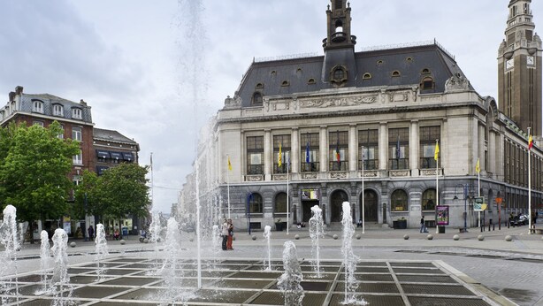 Um edifício histórico com uma fonte em frente, janelas amplas e bandeiras.