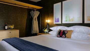 Quarto de hotel com uma cama grande, uma mesinha de cabeceira com garrafa e copos, um roupão pendurado em um cabide e duas fotografias emolduradas na parede.