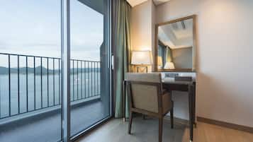 Quarto de hotel com uma grande porta de vidro deslizante que dá para uma varanda com vista para o mar e montanhas. Há uma mesa com cadeira, uma lâmpada e um espelho na parede.
