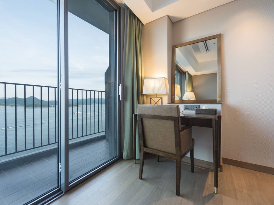 Quarto de hotel com uma grande porta de vidro deslizante que dá para uma varanda com vista para o mar e montanhas. Há uma mesa com cadeira, uma lâmpada e um espelho na parede.