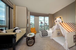 Quarto de hotel com sofá, mesinha redonda, cama com dossel listrado, tenda infantil com tema de panda e uma janela grande com vista para a cidade.