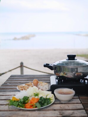 Uma mesa com uma panela, um prato de comida e uma tigela de molho, sobre uma mesa de madeira com vista para a praia ao fundo.