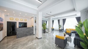 Um hall amplo com balcão de atendimento, vários relógios, área de estar com sofás e uma grande planta.