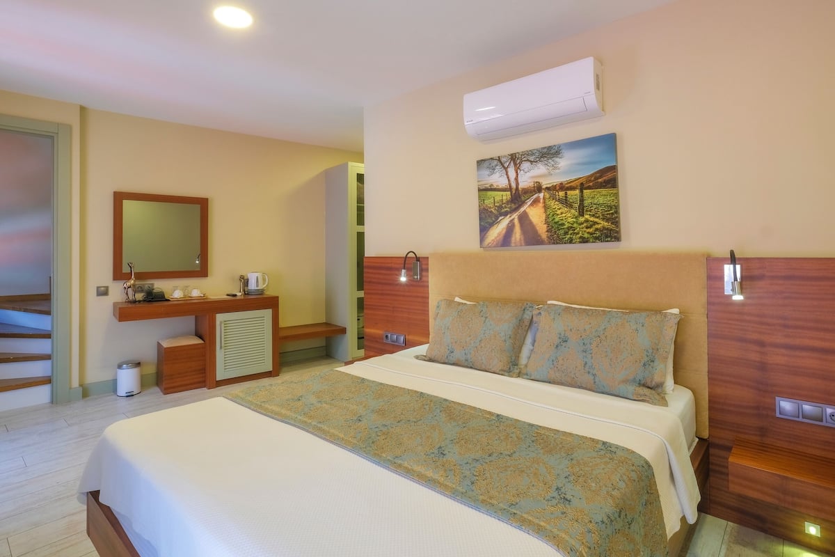 Quarto de hotel com uma cama, uma mesinha de toucador com espelho, um quadro na parede e uma escada.
