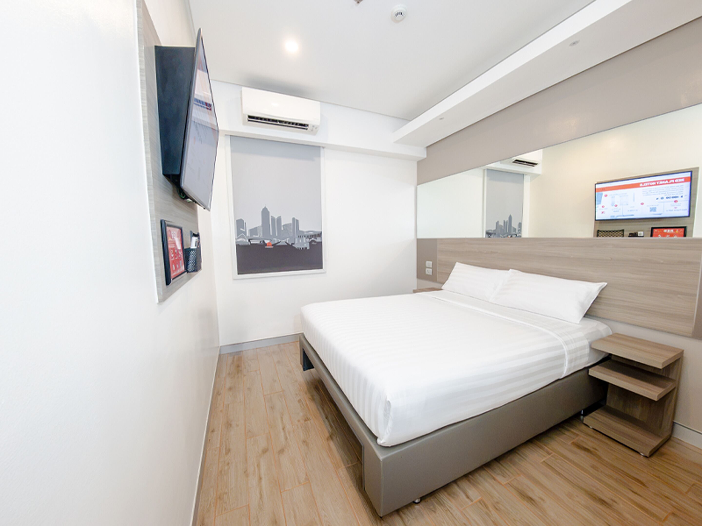 Quarto de hotel moderno com cama, televisão, aparelho de ar condicionado, pôster de paisagem urbana e tela digital.
