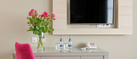 Quarto de hotel com escrivaninha, uma cadeira rosa, um vaso com flores, TV de tela plana na parede e um telefone.