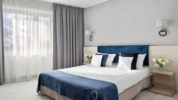 Quarto de hotel com uma cama grande, uma mesa de cabeceira, uma luminária na parede e uma janela com cortinas.