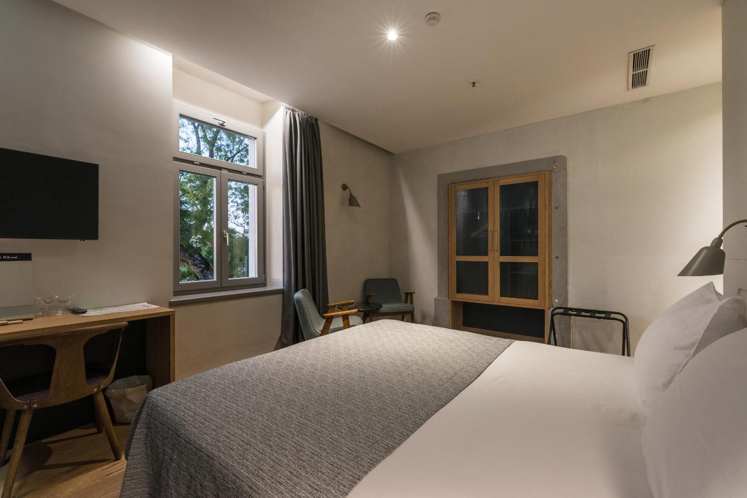 Quarto de hotel com cama, escrivaninha, cadeira, janela e televisão.