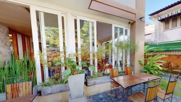 Um pátio externo com mesa e cadeiras de madeira, rodeado por plantas em vasos e piso de pedra.