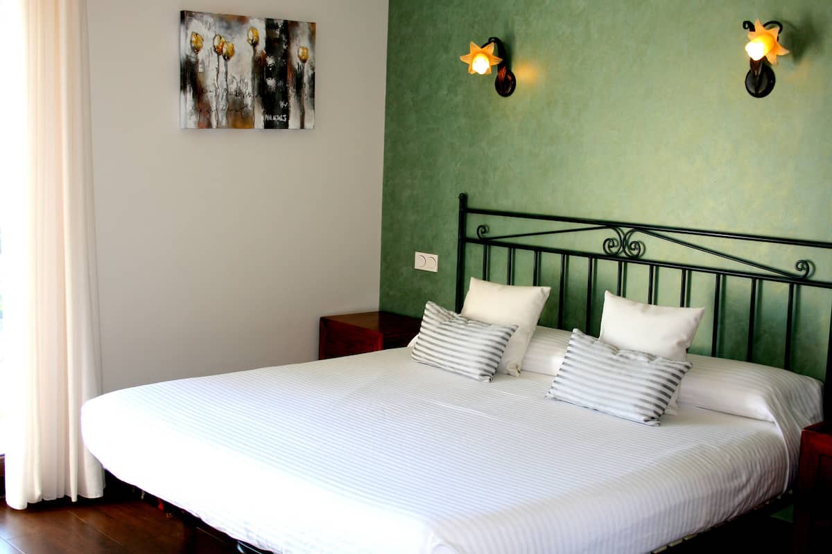 Cama bem arrumada com roupa de cama branca e almofadas listradas, parede verde e duas luminárias fixadas na parede.