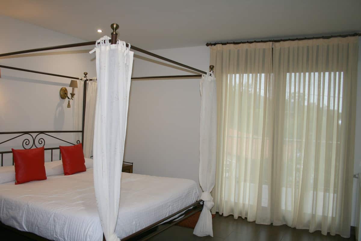 Uma cama com dossel, roupa de cama branca e almofadas vermelhas, uma mesa de cabeceira com abajur e uma janela com cortinas leves.