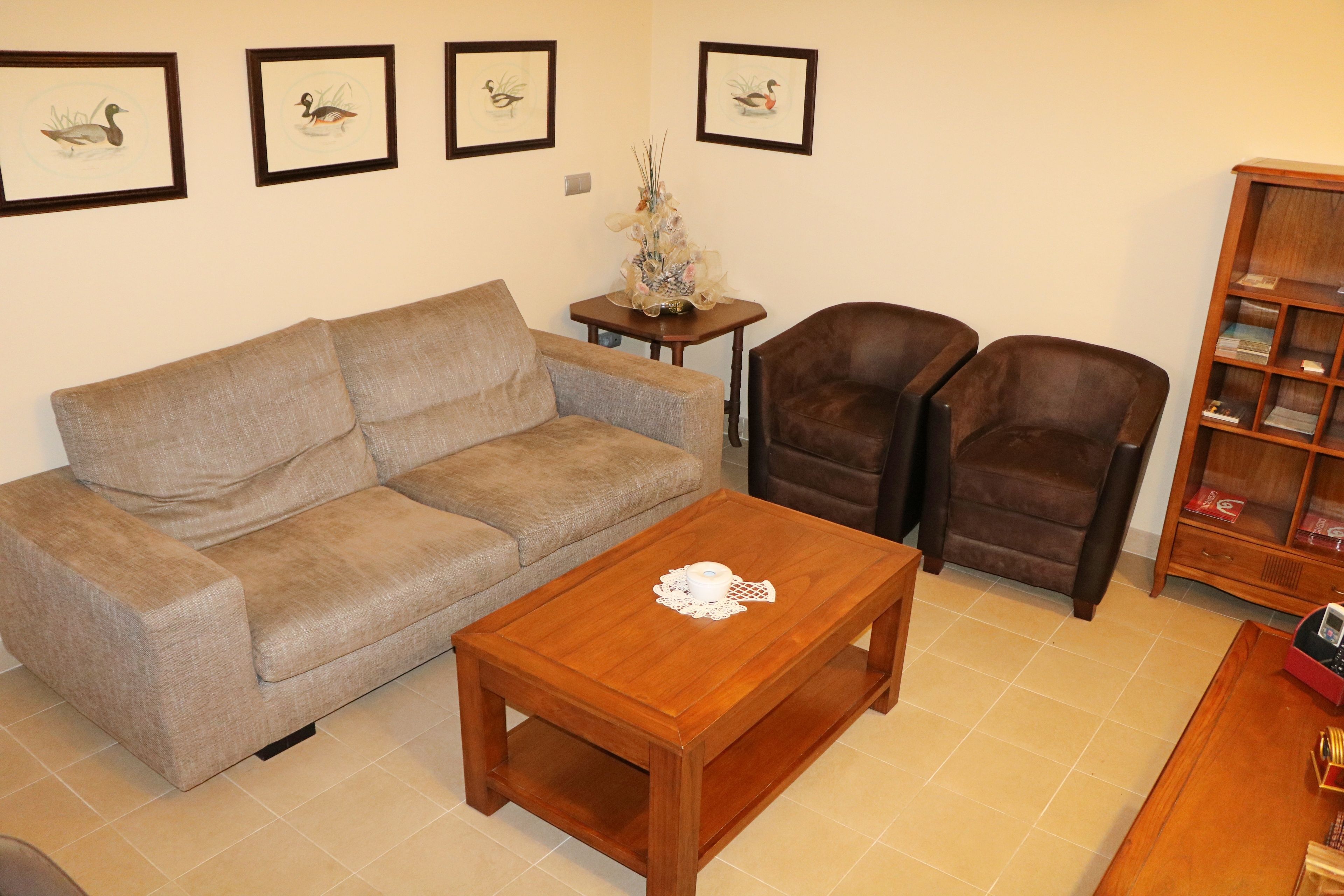 Uma sala de estar com um sofá bege, duas poltronas de couro marrom, uma mesa de centro de madeira e uma estante com livros e objetos decorativos.