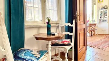Quarto com cadeira e mesa brancas, janela com cortinas azuis e porta que dá para outro cômodo.