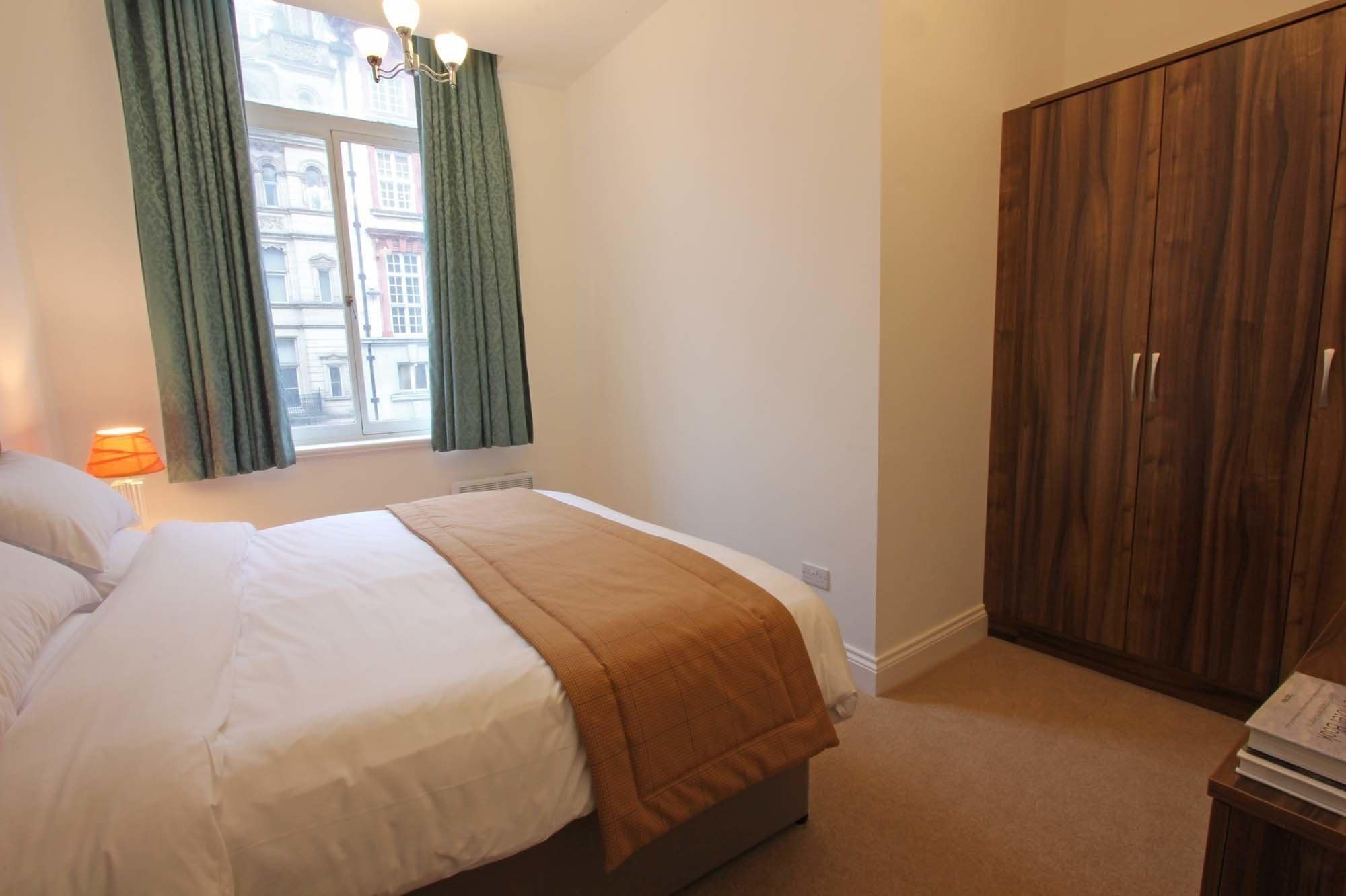 Um quarto com cama, janela com cortinas, abajur e guarda-roupa.