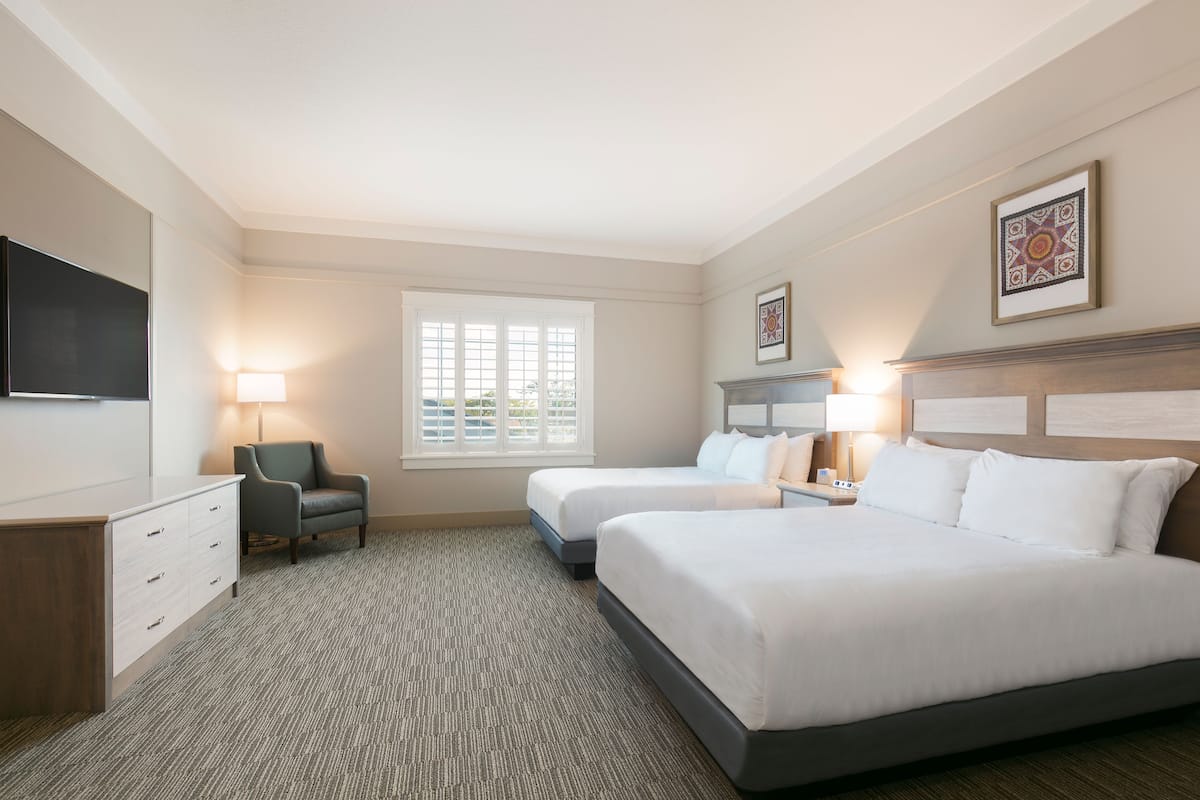 Quarto de hotel com duas camas, uma escrivaninha, uma cadeira, uma televisão e uma janela com persianas.