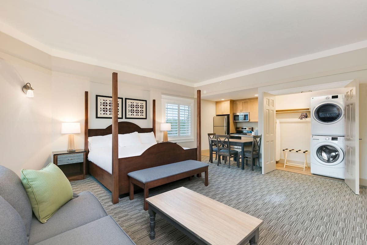 Quarto de hotel com cama de dossel, sofá, mesa de jantar com cadeiras, área de cozinha com eletrodomésticos e máquina de lavar roupa.