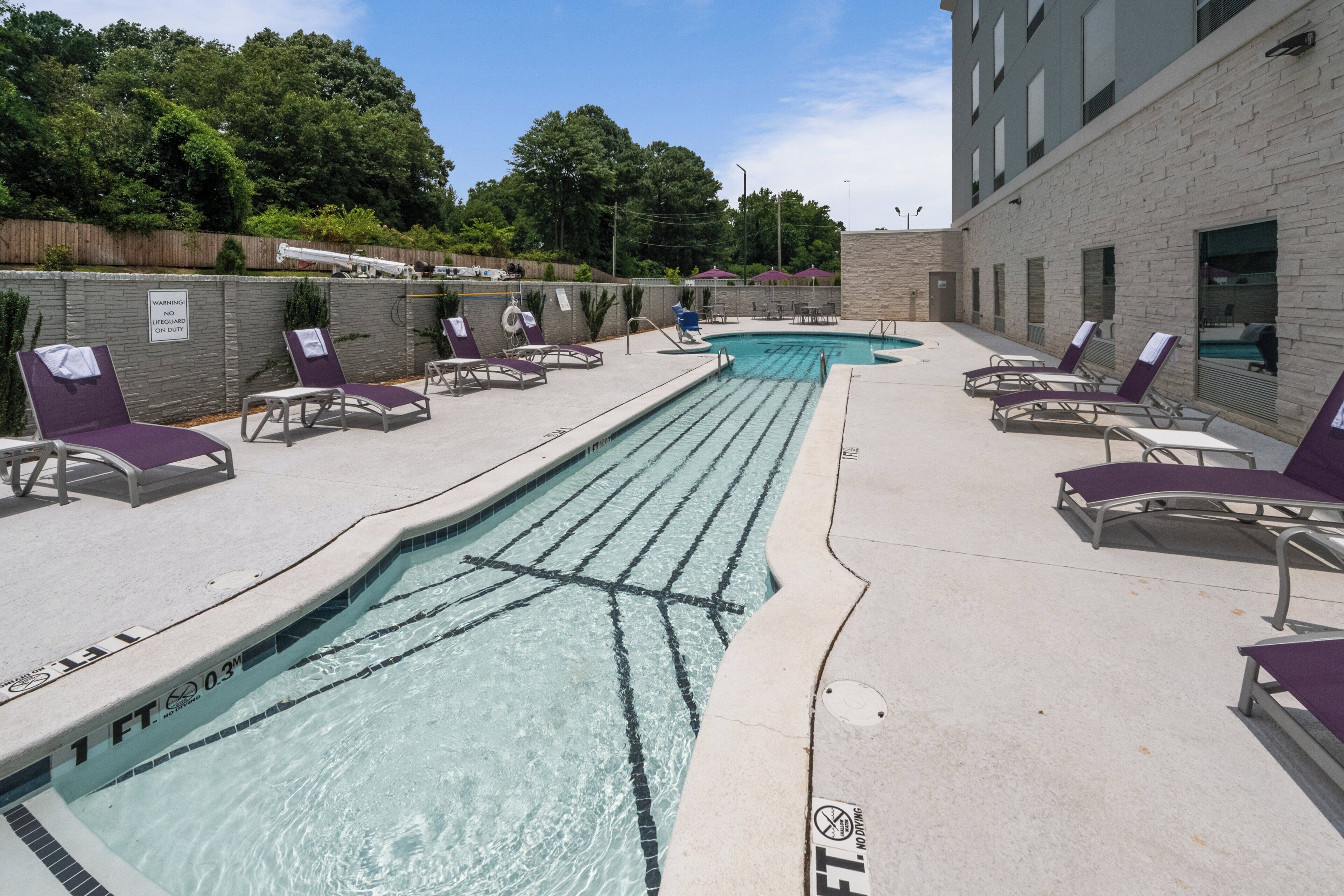 Piscina com várias pistas, rodeada por espreguiçadeiras e um edifício de tijolo.