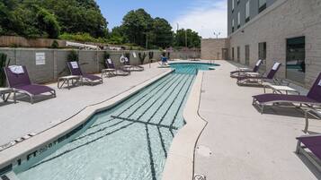 Piscina com várias pistas, rodeada por espreguiçadeiras e um edifício de tijolo.