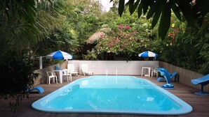 Piscina retangular com um deque de madeira, rodeada por espreguiçadeiras brancas e dois guarda-sóis azuis.