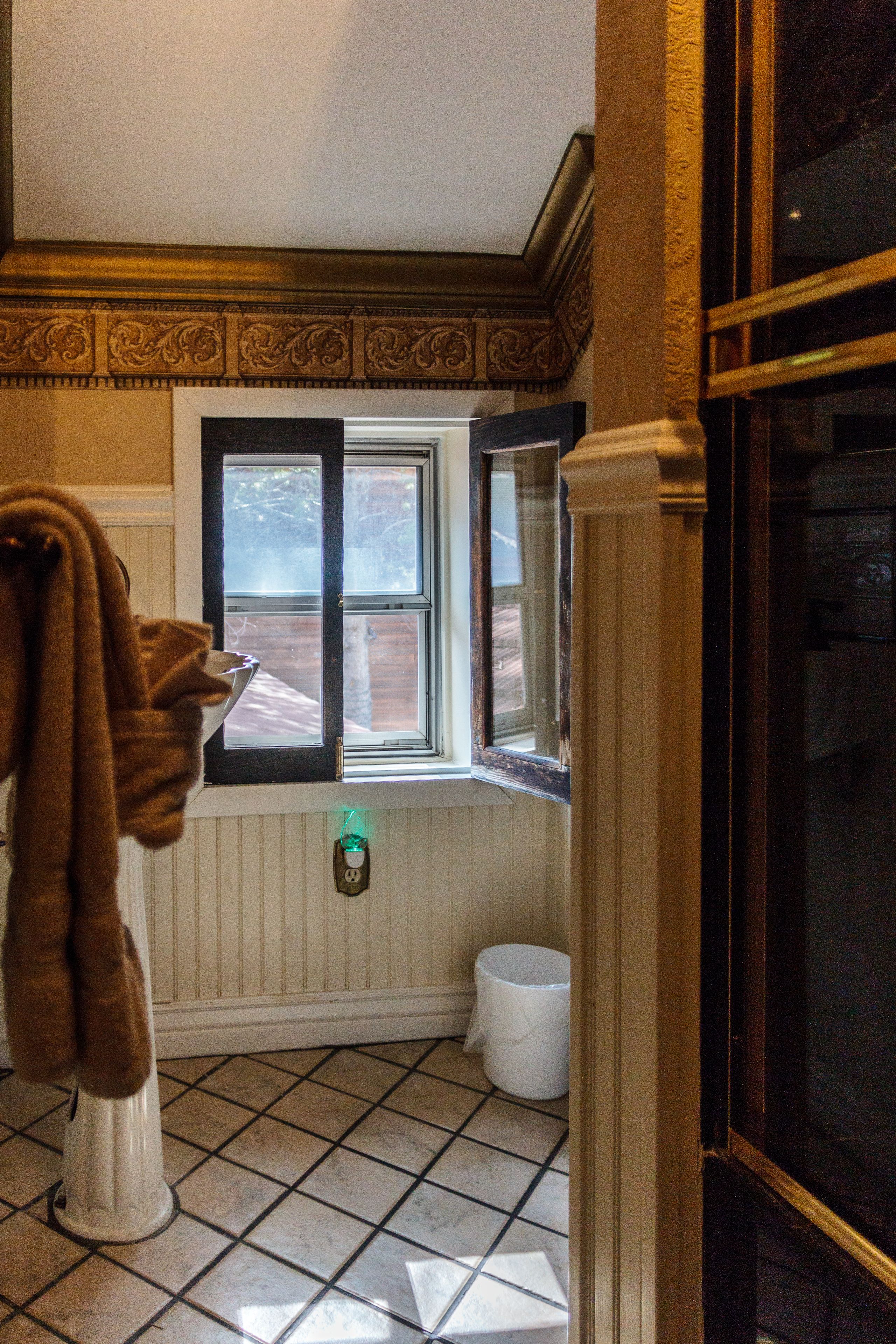 Casa de banho com janela, um porta-toalhas, uma sanita pedestal branca e um balde branco.