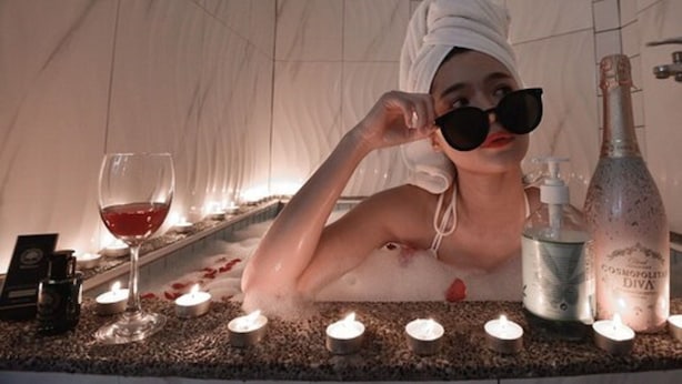 Uma pessoa em banheira, com velas, uma garrafa e um copo com líquido vermelho.
