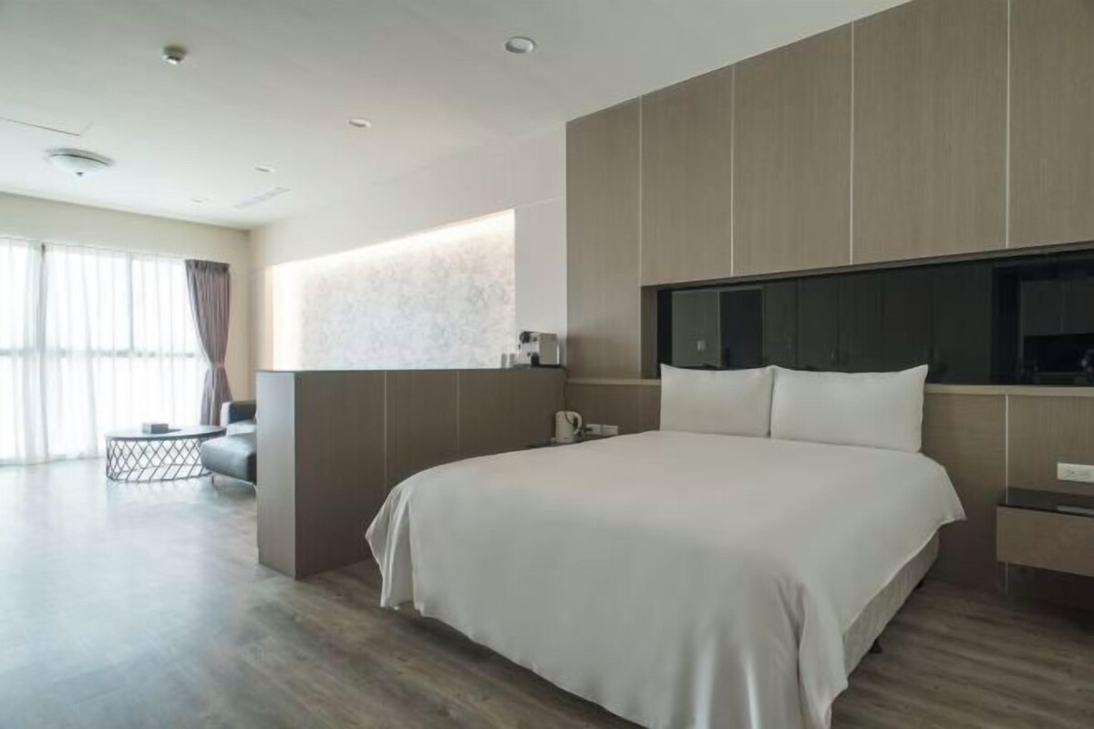 Quarto de hotel moderno com uma cama grande, televisão integrada, mesa de cabeceira e uma janela ampla com cortinas.