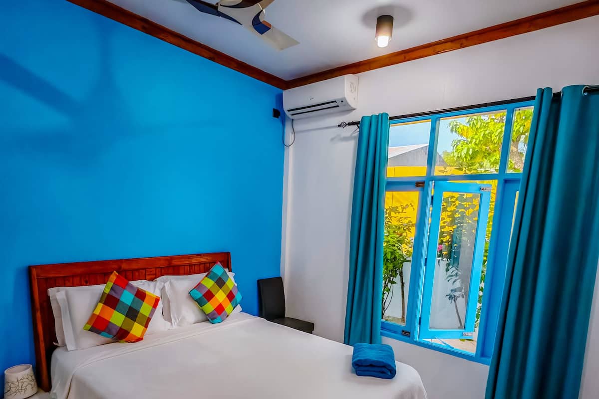 Quarto de hotel com cama, ventoinha no teto, ar condicionado, janela com cortinas e vista para o verde lá fora.
