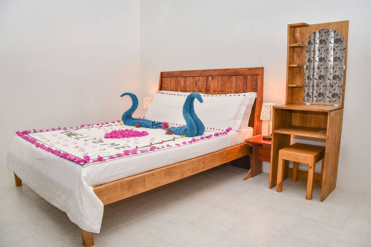 Uma cama com cabeceira de madeira, mesa de cabeceira, criado-mudo de madeira e um pequeno banquinho de madeira.