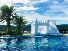 Um castelo inflável junto a uma piscina, com palmeiras e vista para a montanha.