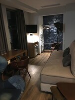 Uma sala de estar moderna com um sofá, uma escrivaninha e uma cadeira. Há uma TV fixada na parede e uma janela com cortinas.