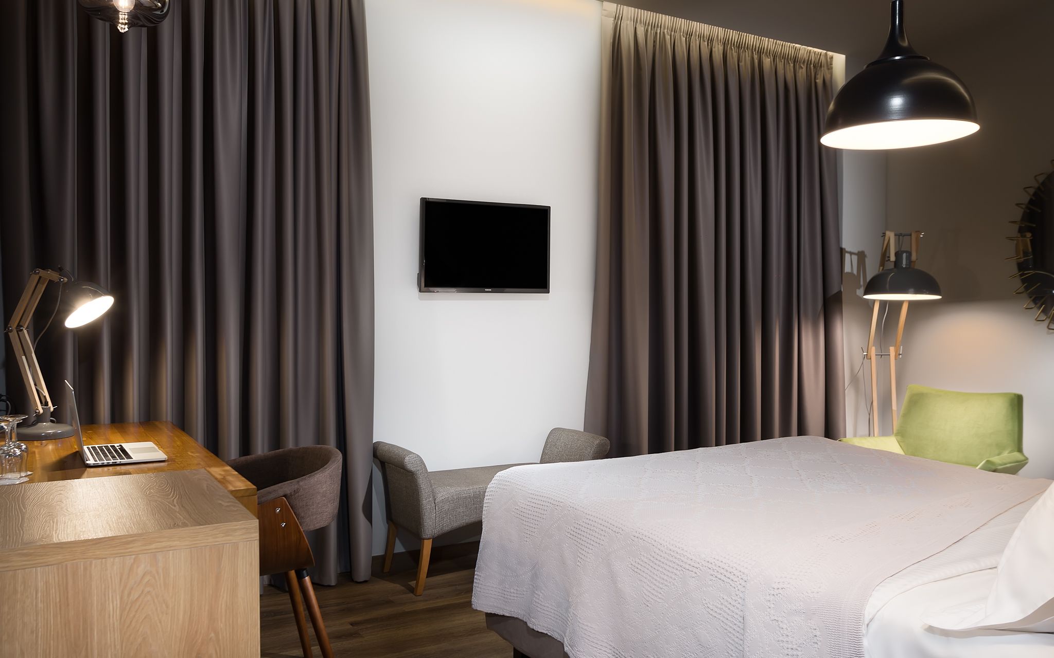 Quarto de hotel com cama, secretária com um laptop, uma cadeira, televisão e duas luminárias.