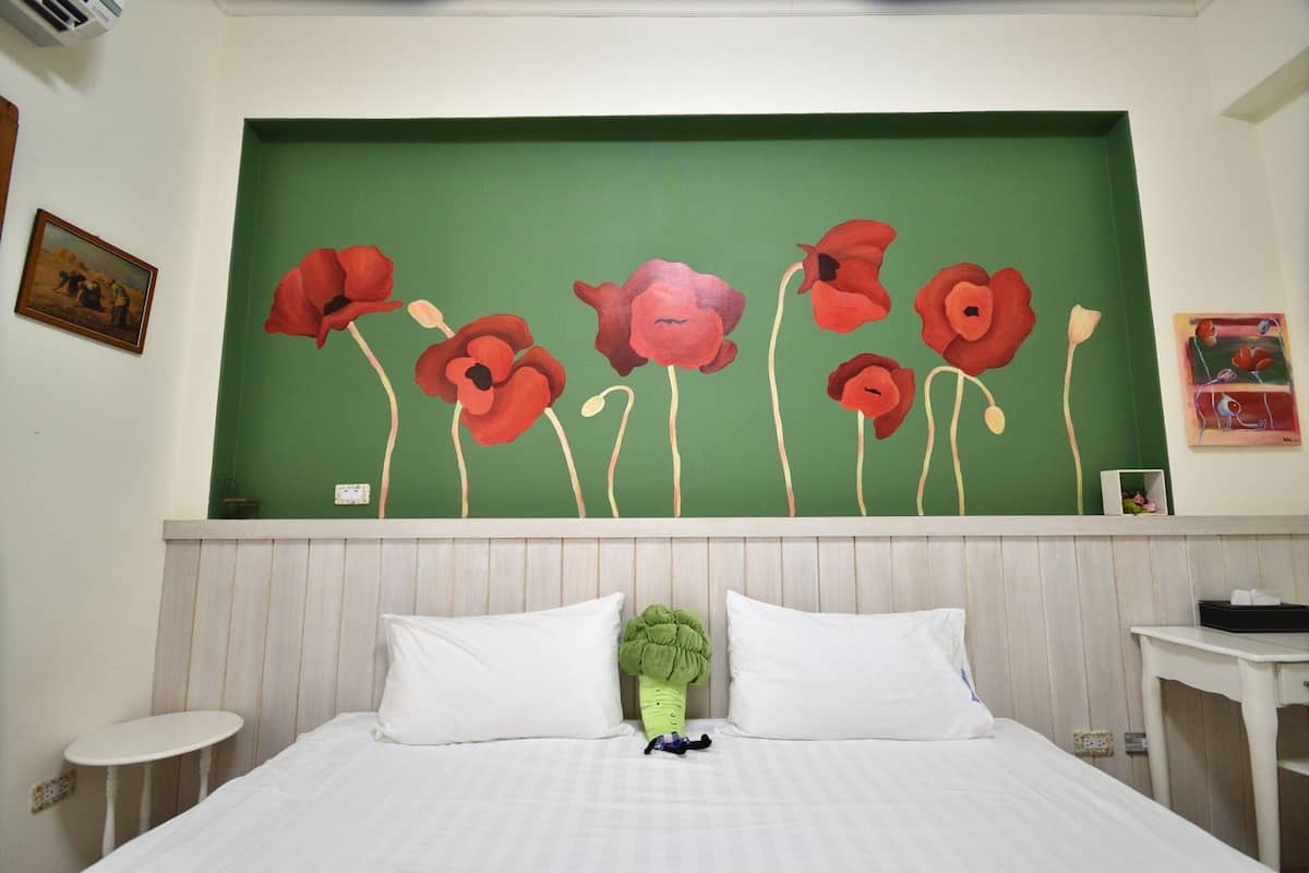 Quarto de hotel com cama, duas almofadas, parede verde com pintura de papoula, mesa redonda pequena e mesa de cabeceira.