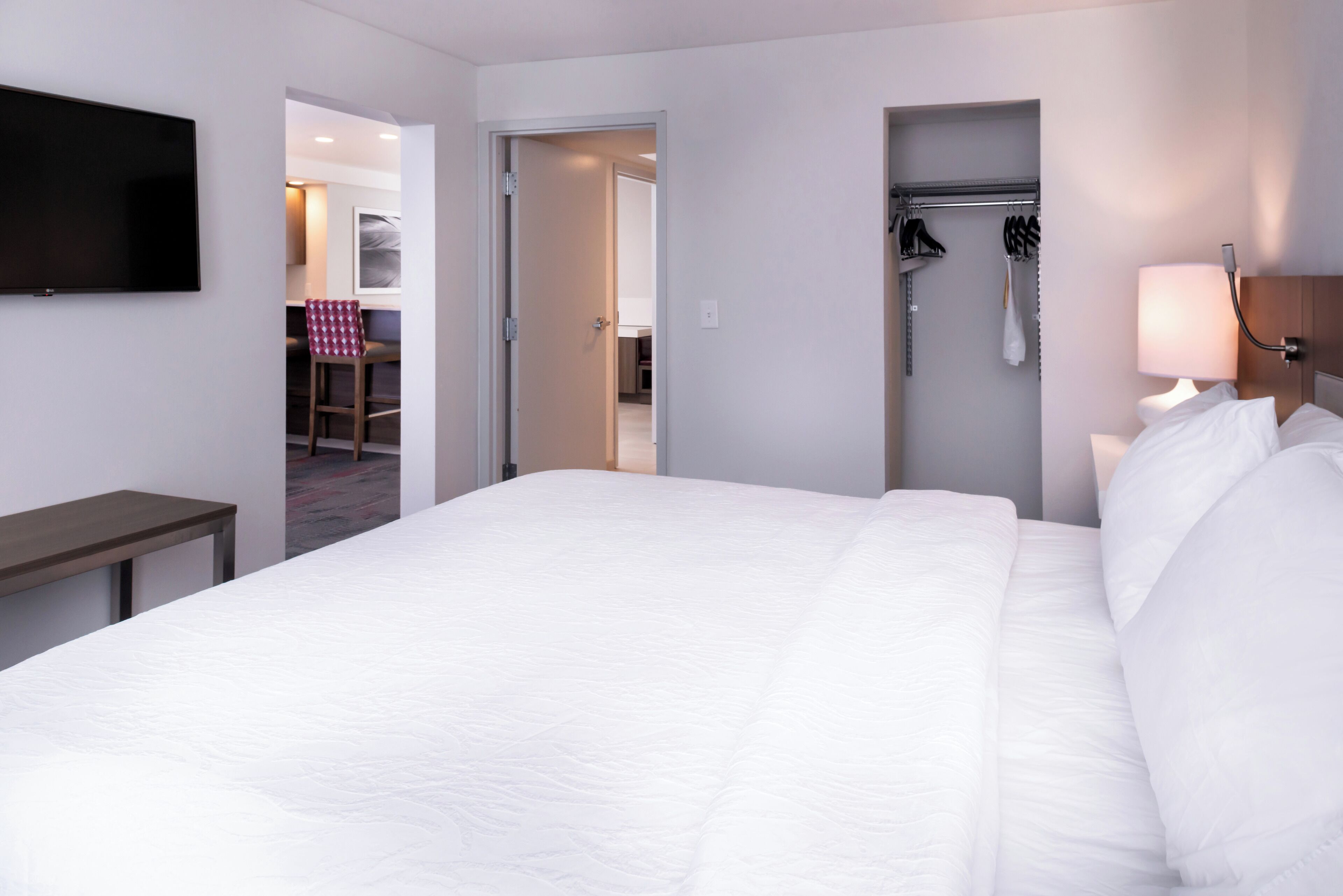 Quarto de hotel com uma cama grande, televisão na parede, escrivaninha e closet espaçoso com espelho e cabideiro.