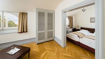 Quarto com piso de madeira em padrão herringbone, cama com roupa de cama branca, mesa de cabeceira, janela com cortinas e porta com mosquiteiro.