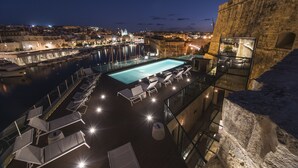 Piscina infinita no terraço com espregui çadeiras, com vista para um porto com barcos e luzes da cidade.