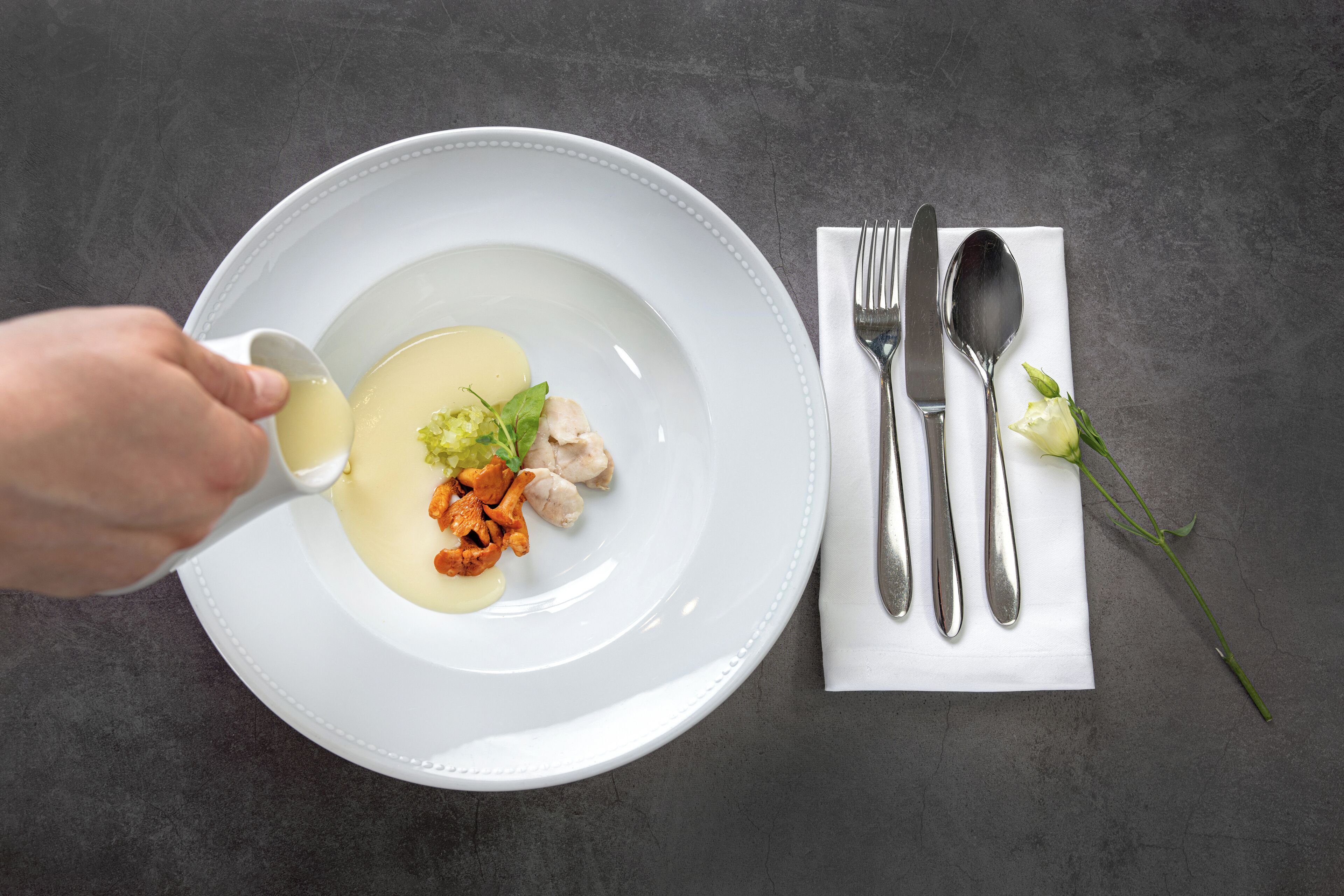 Um prato branco com sopa e guarnições, uma colher, garfo e faca sobre um guardanapo, e uma mão segurando uma colher.