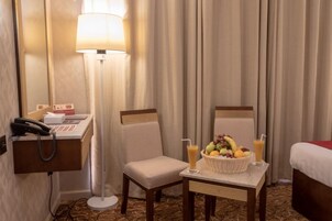 Quarto de hotel com escrivaninha, telefone, cadeira, mesa com cesta de frutas e bebidas, e uma cama.