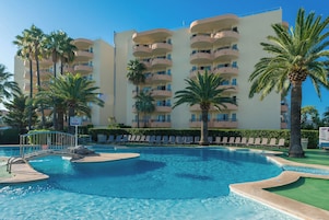 Um hotel com várias varandas, piscina com espreguiçadeiras e palmeiras.