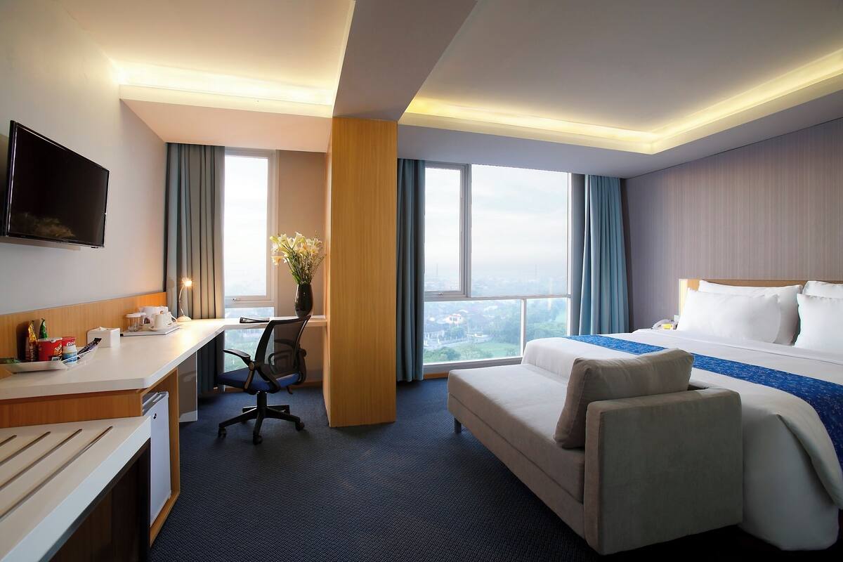 Quarto de hotel com cama, escrivaninha, cadeira, televisão e uma janela grande.